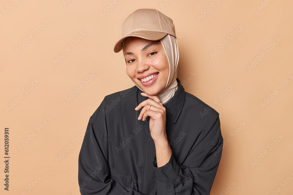 Portrait of positive Muslim woman keeps hand under chin smiles gently ...