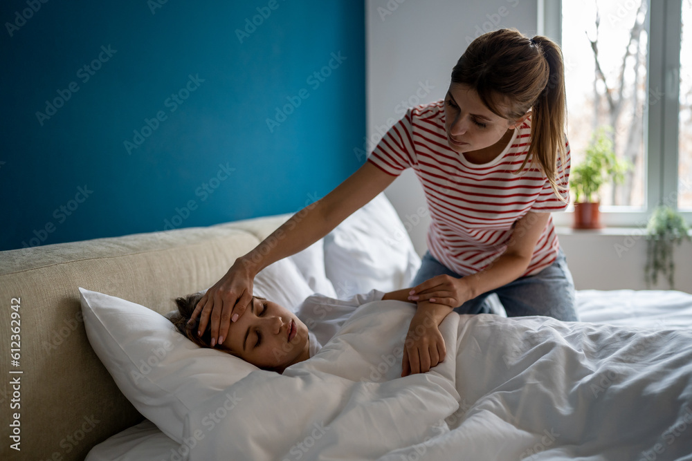 Sick daughter lying in bed, having chills and fever. Mother checking temperature touching ...