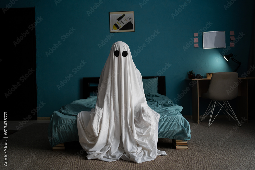 Ghost in white sheet sits on bed in bedroom at night. Horror scene ...