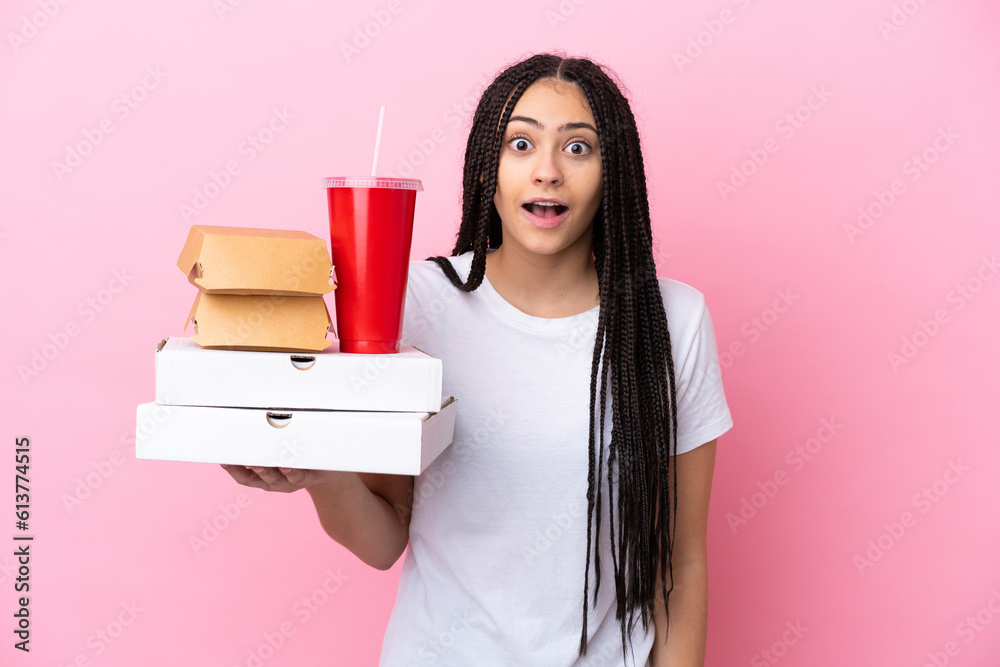Teenager girl with braids holding pizzas and burgers over isolated pink ...