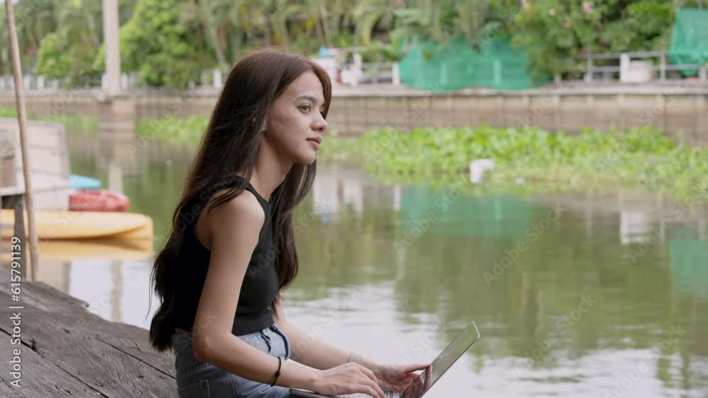 Asian woman working by the canal, sits with legs dangling laptop ...