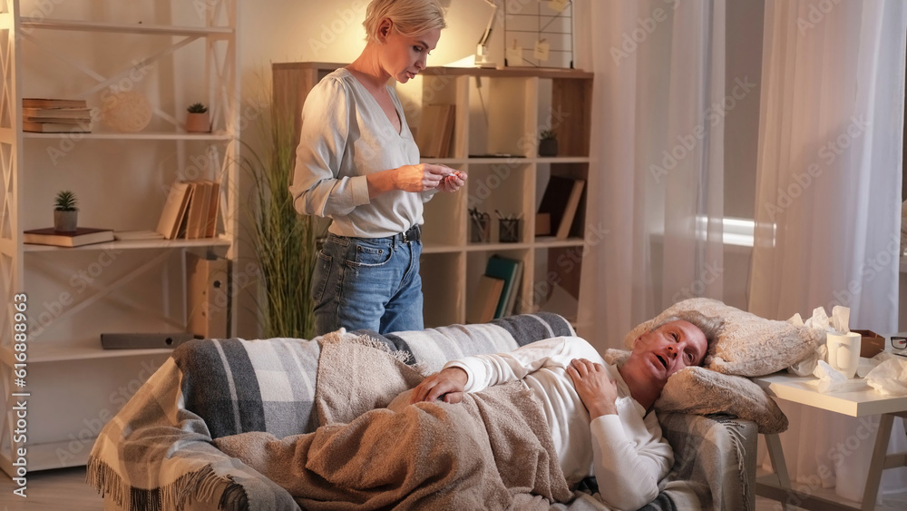 Caring husband flu. Fever care. Woman taking temperature sick man laying on sofa under blanket ...