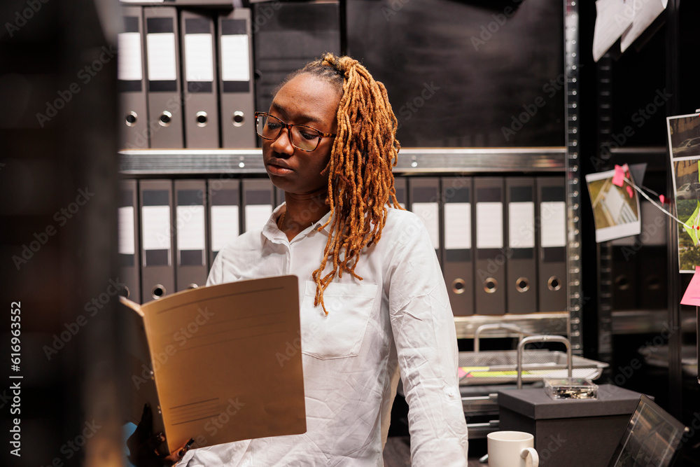 Policewoman standing near crime case folders shelf and reading report ...