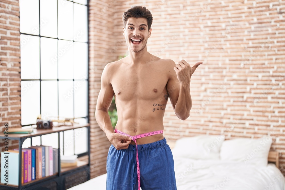 Young hispanic man using tape measure measuring waist pointing thumb up ...