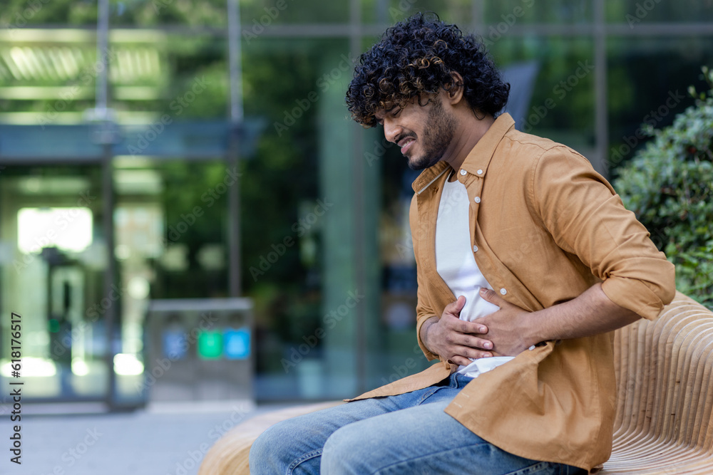 A young Indian man sits outside on a bench and suffers from stomach ...
