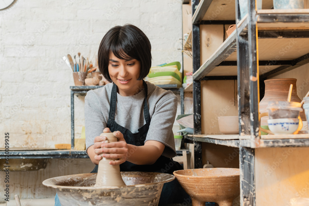 Smiling young asian female artist in apron molding clay on pottery ...