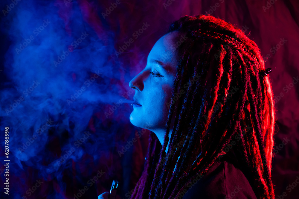 Profile of caucasian girl with dreadlocks smokes a vape in red blue ...