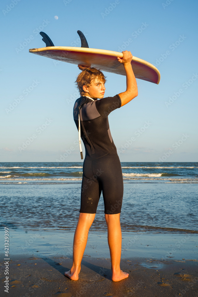 Teenage surfer boy standing on the beach in wetsuit holding his ...
