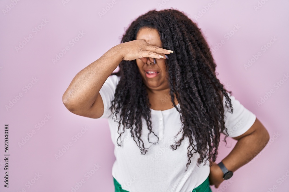 Plus size hispanic woman standing over pink background smelling ...