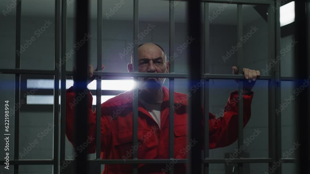 Elderly prisoner in orange uniform holds hands on metal bars. Criminal ...