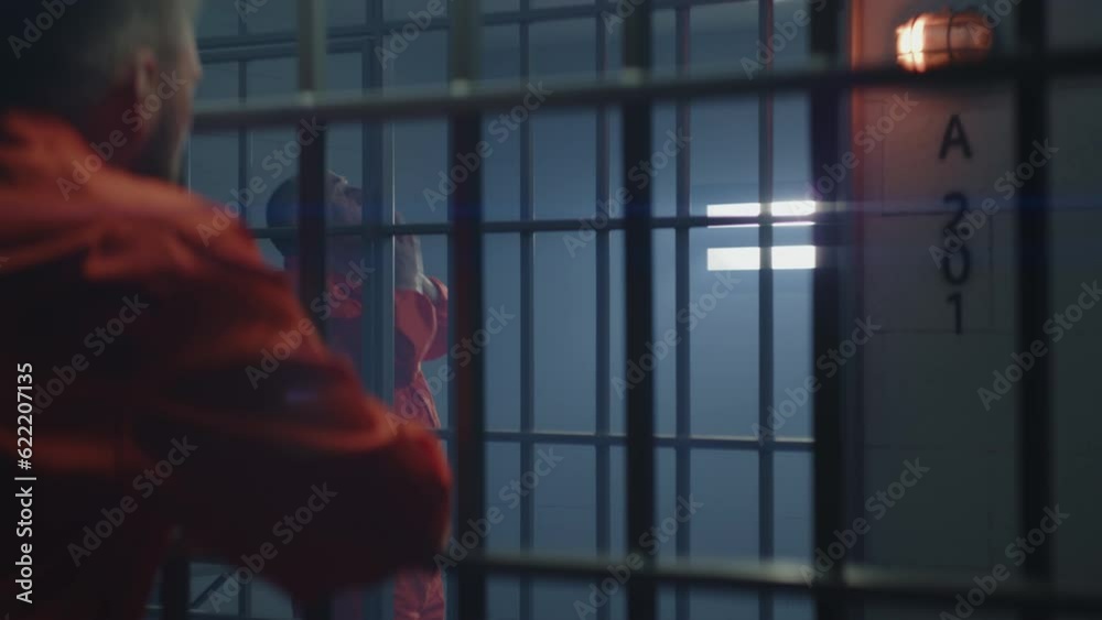 Man in orange uniform stands in jail cell and holds hands on bars ...