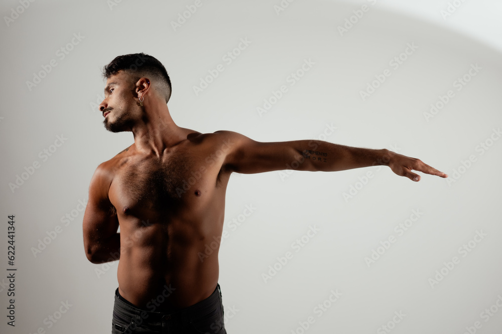 A shirtless man in a white studio stands tall and flexes his body, with ...