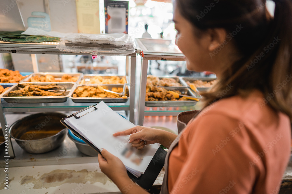 female stall owner checks the ready menu using a paper checklist on a ...