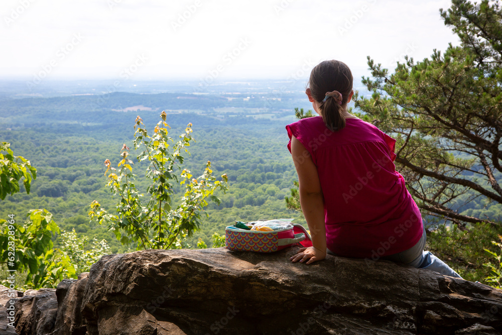Young girl sitting and resting on cliff rock with picnic lunch looking ...