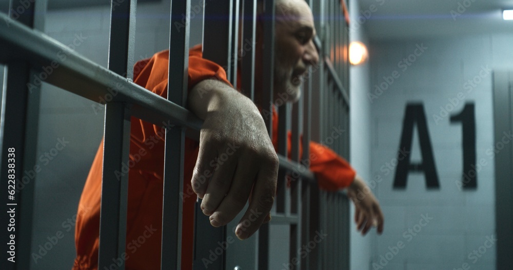 Elderly prisoner in orange uniform stretches his hands, leans on metal ...