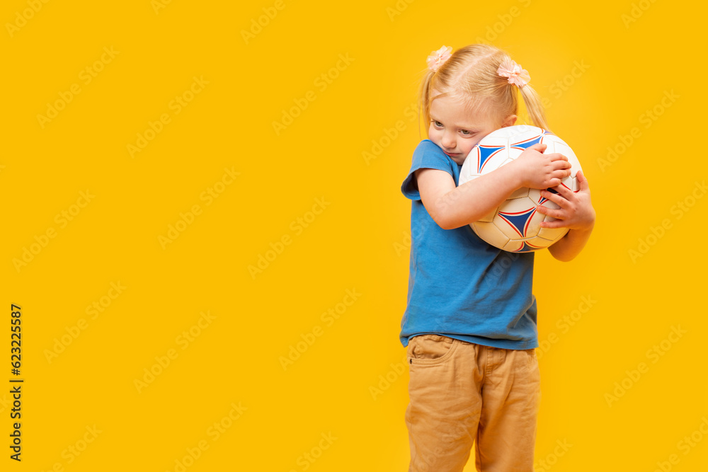Little blonde girl hugs soccer ball. yellow studio background. Copy ...