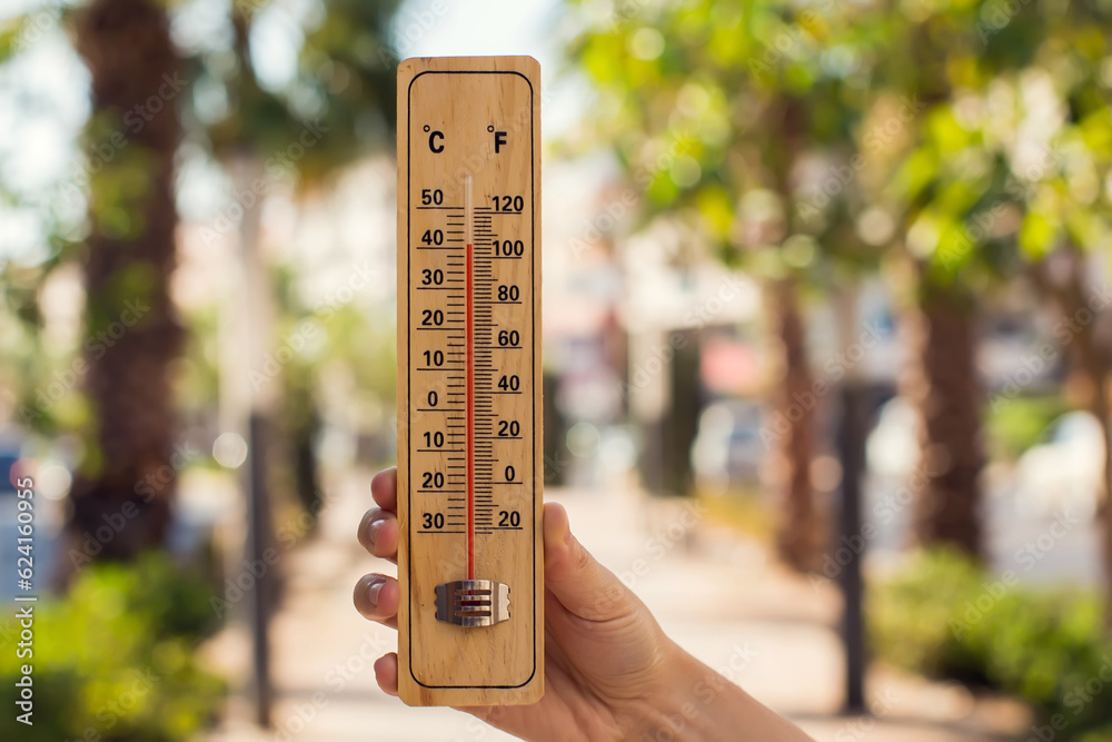 Hot weather. Thermometer in hand in front of street with trees and ...