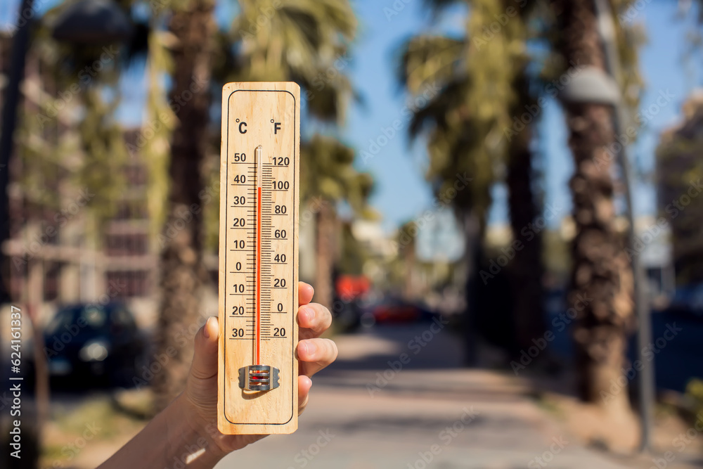 Hot weather. Thermometer in hand in front of street with trees and ...