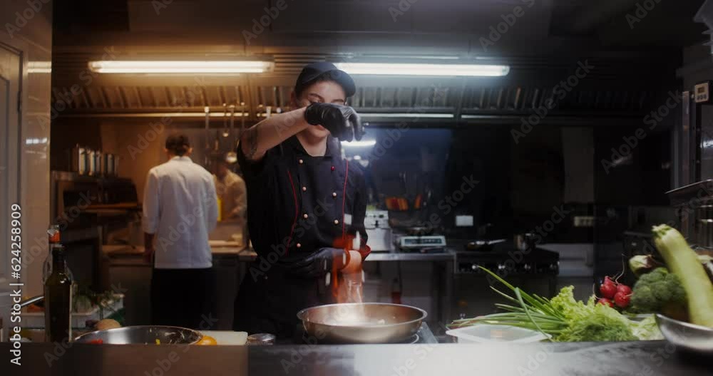 A woman in a chef's uniform adds food to a frying pan with fire while ...