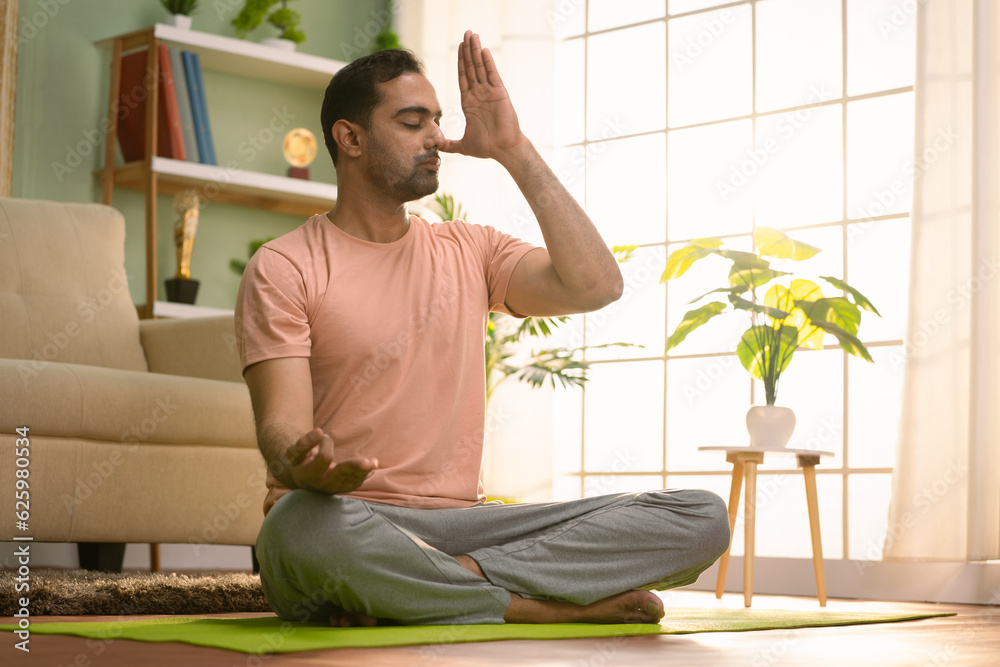 Peaceful indian man doing nostril breathing or pranayama yoga by closing eyes during morning at ...