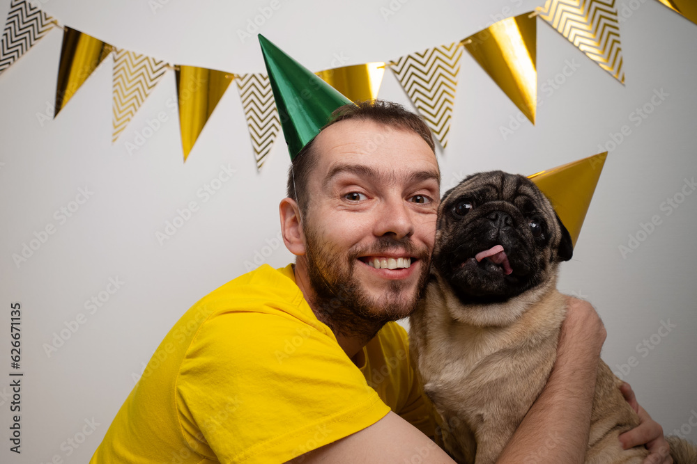 Funny guy with cute pug dog in a party hat hugging and celebrating pet ...