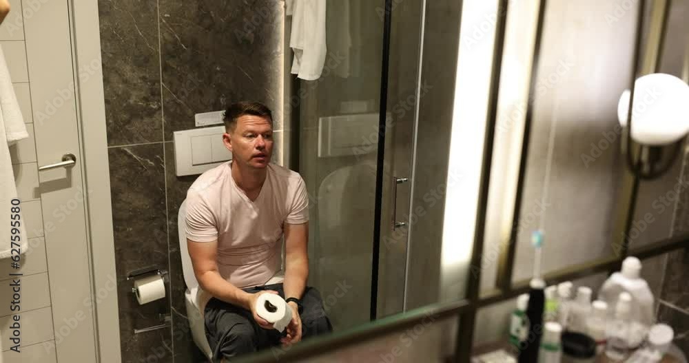 Young man sits at home on toilet bored. Sad man sits on toilet with ...