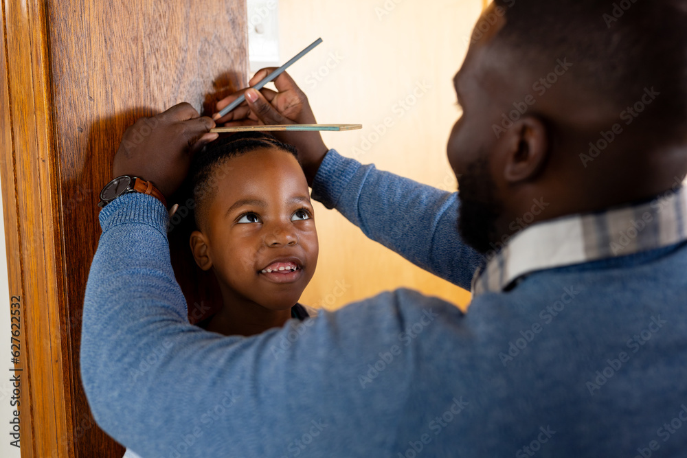 Happy african american father measuring son and marking height on door ...