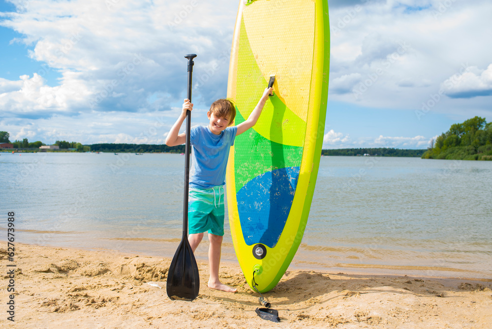 happy little boy stands with his green supboard and paddle on the beach ...