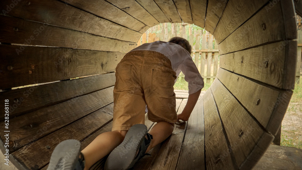 Cute boy crawling through wooden tube or tunnel at summer adventure ...