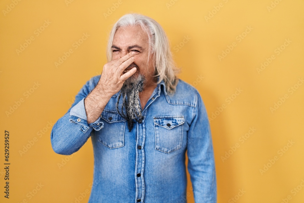 Middle age man with grey hair standing over yellow background smelling ...