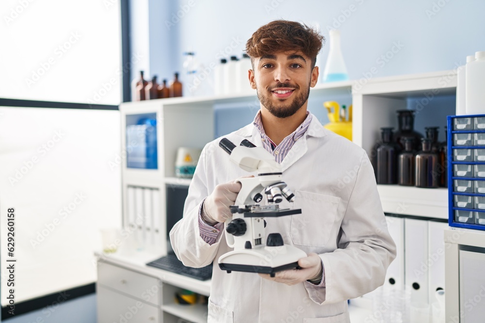 Arab man with beard working at scientist laboratory holding microscope smiling with a happy and ...
