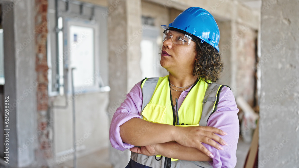 Young beautiful latin woman builder standing with serious face and arms ...