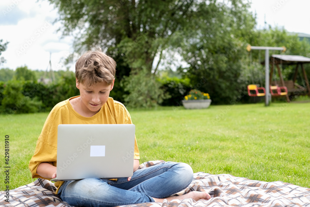 TTeen boy with a laptop sits in a park. School boy is typing on ...