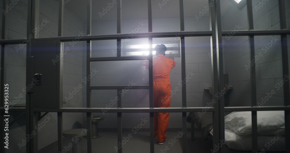African American prisoner in orange uniform looks at barred window and ...