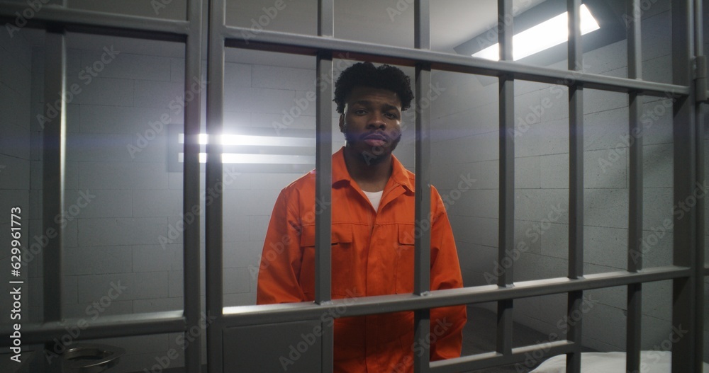 African American man in orange uniform leans hands on prison cell bars ...