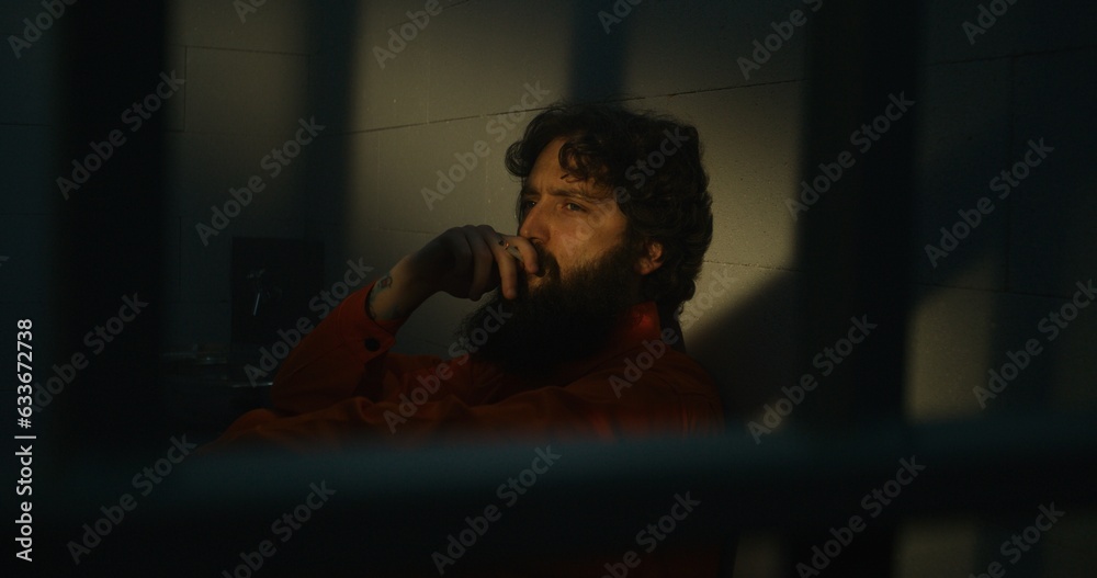 Depressed prisoner in orange uniform sits on bed, smokes cigarette in ...