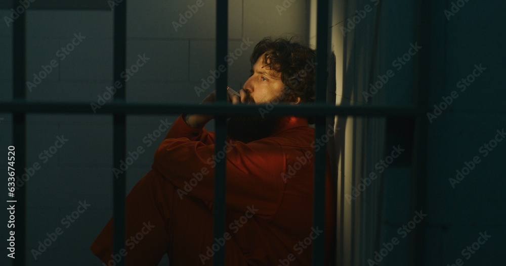 Pensive male prisoner in orange uniform sits on bed, smokes cigarette ...