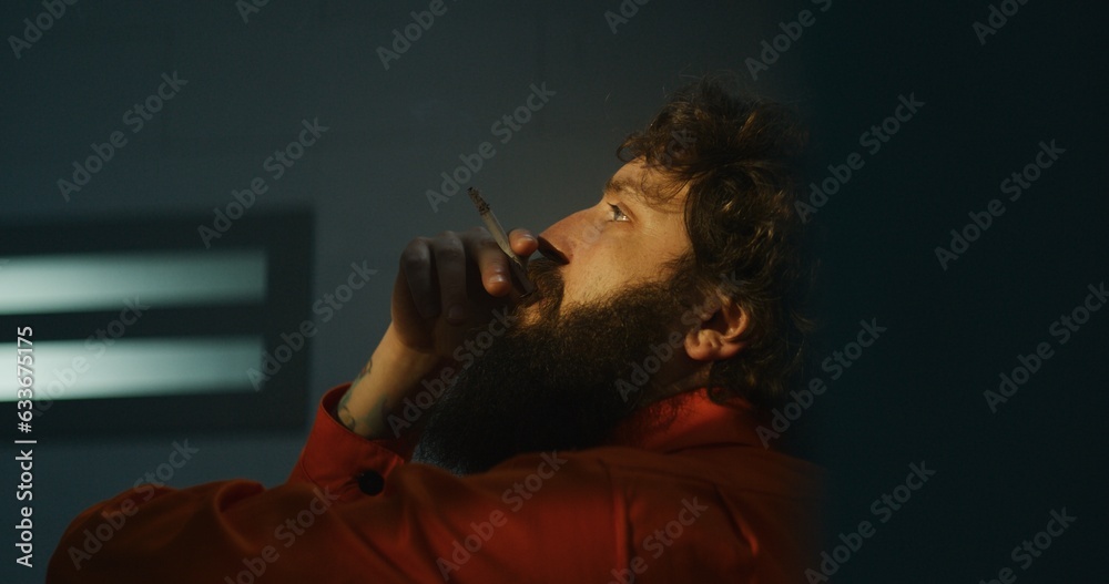 Pensive male prisoner in orange uniform sits on bed, smokes cigarette ...