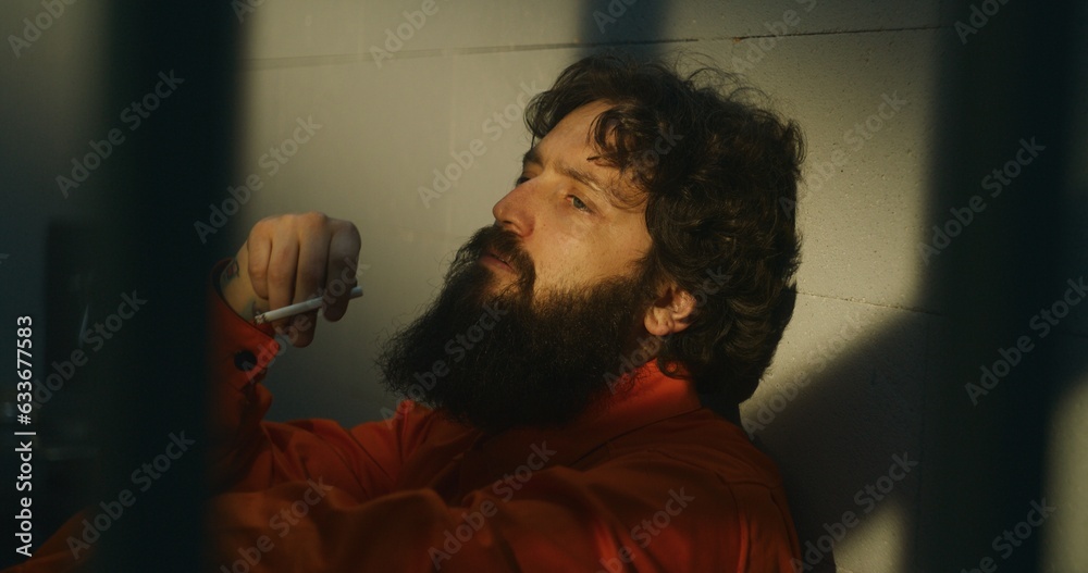 Thoughtful male prisoner in orange uniform sits on the bed, lights ...