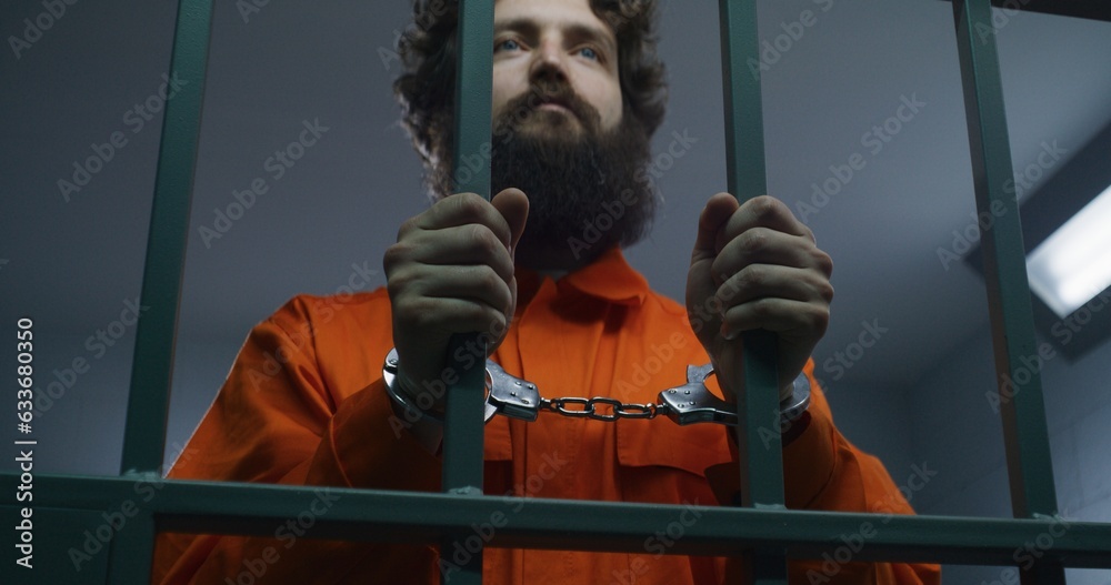 Male prisoner in orange uniform holds metal bars, stands in prison cell ...