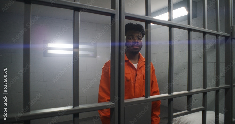 African American man in orange uniform leans hands on prison cell bars ...