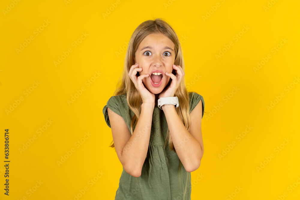 Stupefied blonde kid girl wearing green T-shirt over yellow studio ...