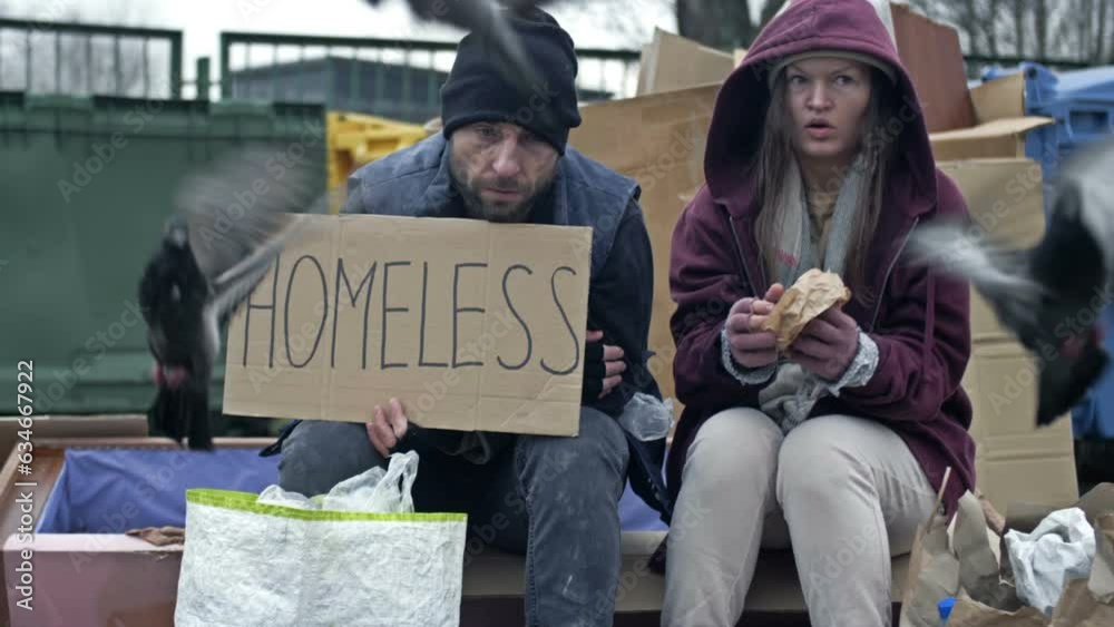 Two dirty and poorly dressed homeless people, a man and a woman, sit by ...
