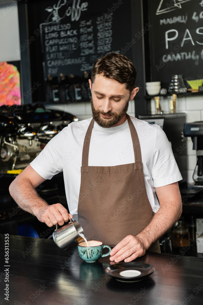 bearded barista in apron pouring milk from pitcher while making ...
