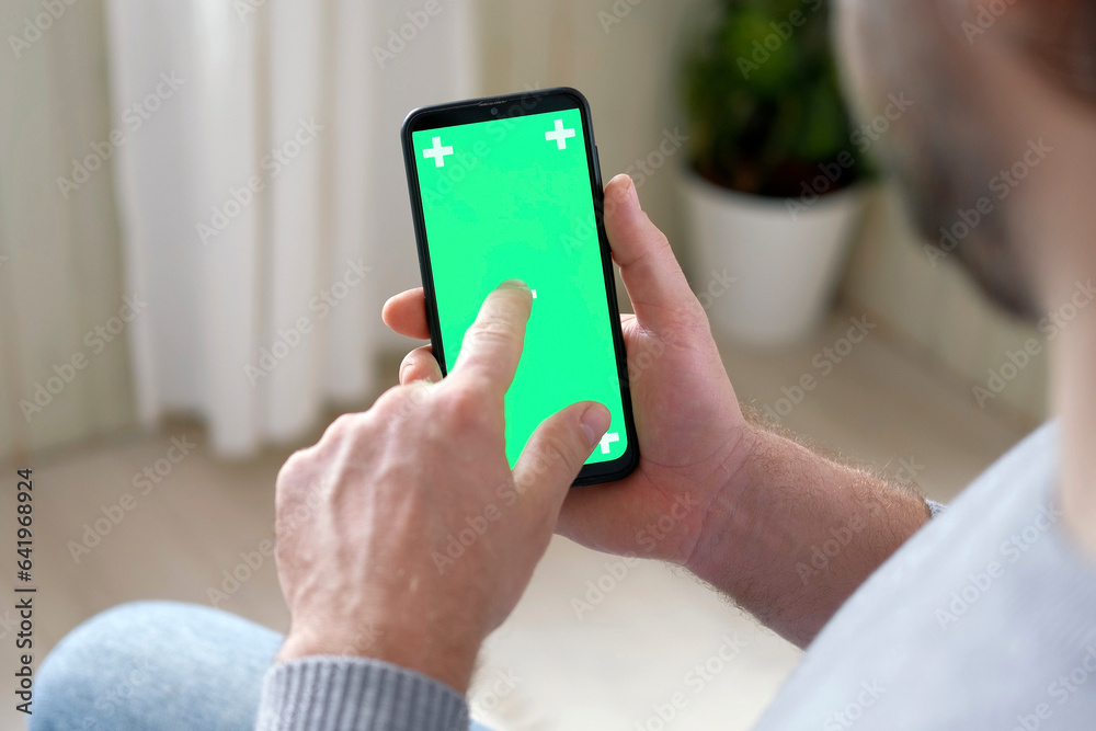 Young man sitting at home holding smartphone green mock-up screen in hand. Male person using ...
