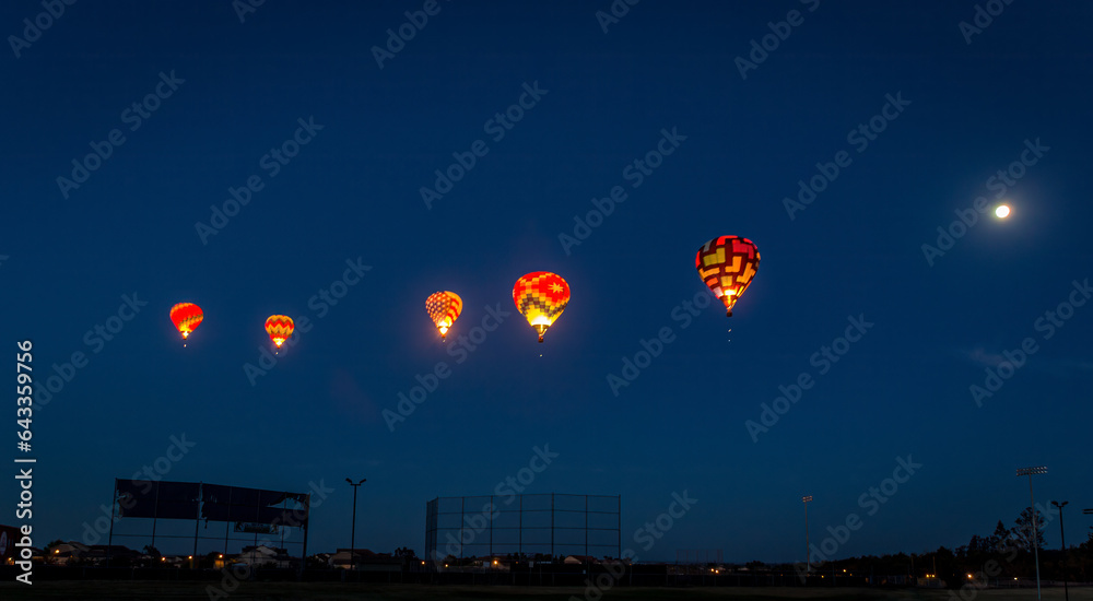 Five hot air balloons are flying over a baseball field at dawn. All ...
