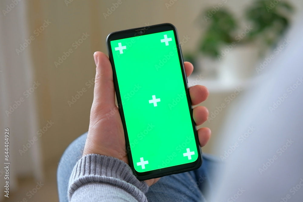 Young man sitting at home holding smartphone green mock-up screen in ...