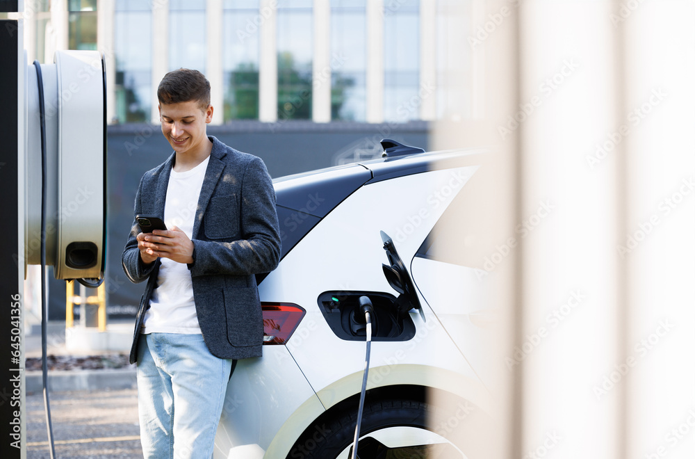 Caucasian man using smartphone and waiting power supply connect to electric vehicles for ...