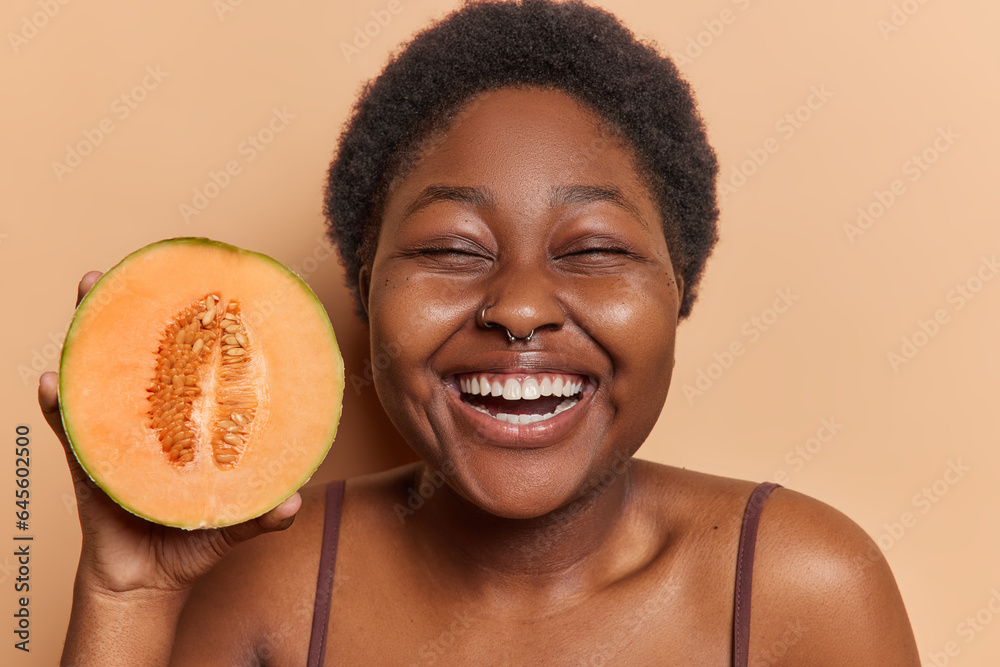 Overjoyed dark skinned woman smiles happily holds half of fresh melon ...