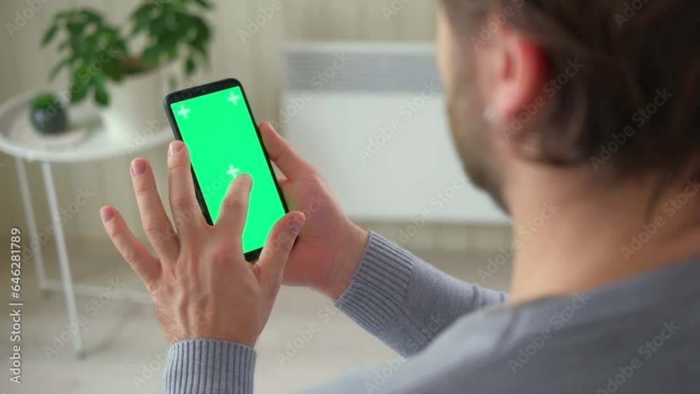 Young man sitting at home holding smartphone greenscreen mock-up in ...
